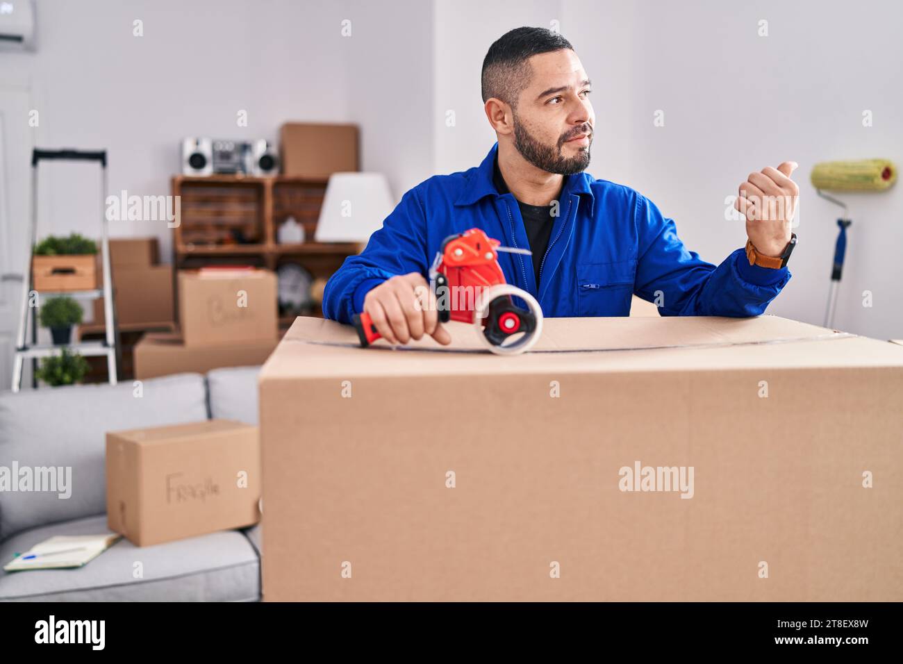 Hispanic man working on moving holding packing tape pointing thumb up ...