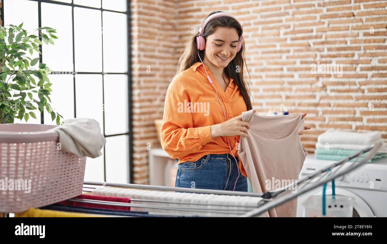Young beautiful hispanic woman listening to music hanging clothes on ...