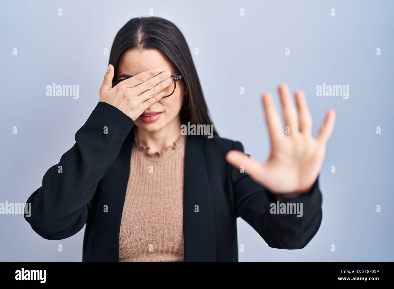 Young brunette woman standing over blue background covering eyes with ...