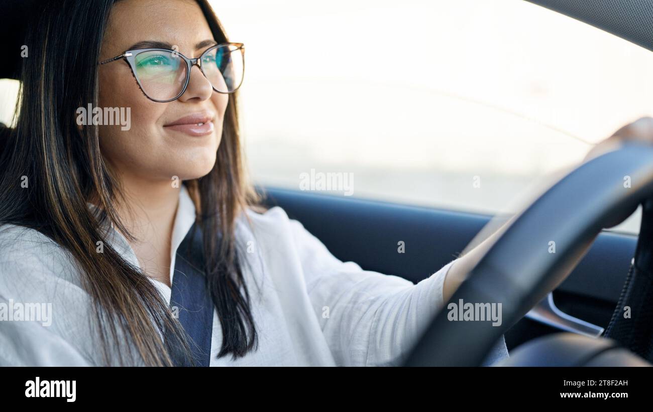 Young beautiful hispanic woman driving a car smiling wearing glasses on ...