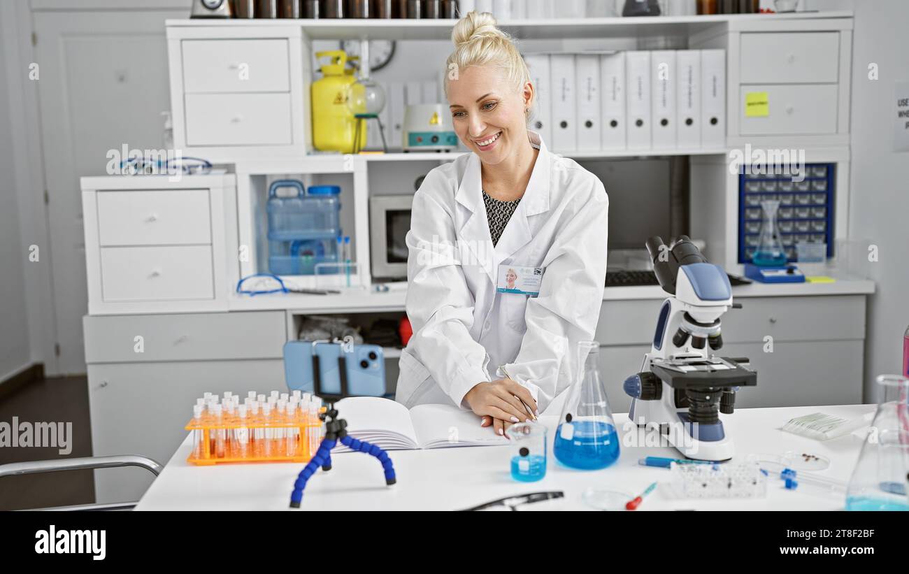 Charming, blonde scientist engrossed in note-taking during video call ...