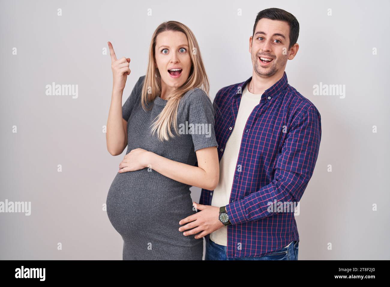 Young couple expecting a baby standing over white background pointing ...