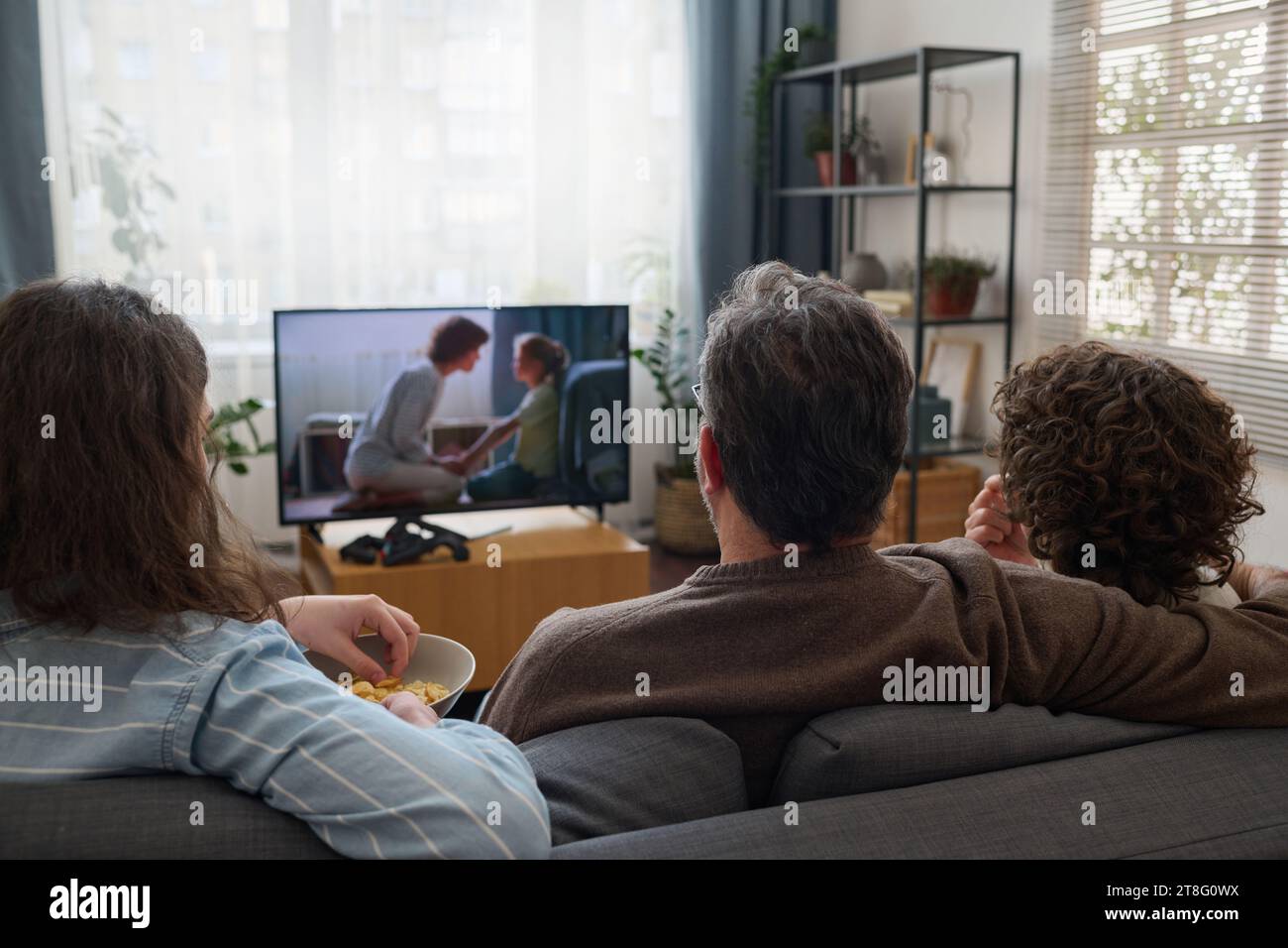Rear view of family watching TV together in the living room while ...