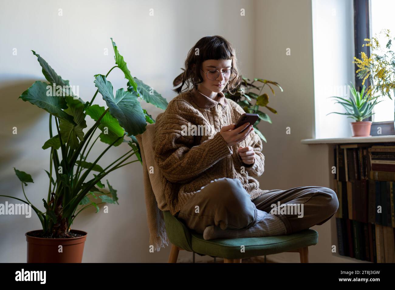 Calm teen girl in glasses looking in smartphone screen sitting on chair ...