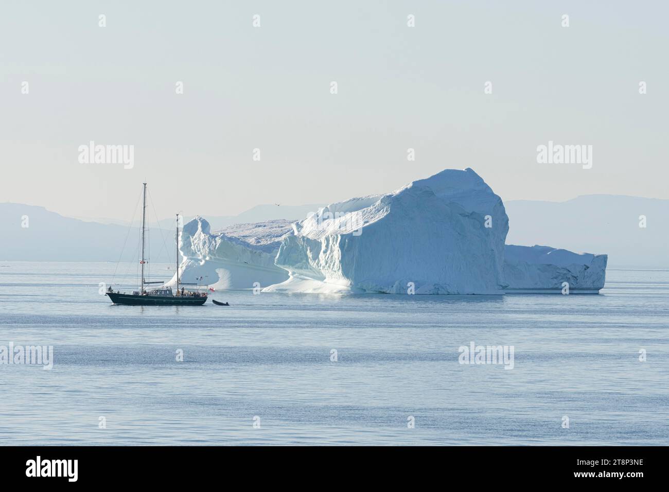 A small sailing ship passes an iceberg in the evening sun. Disko Bay ...