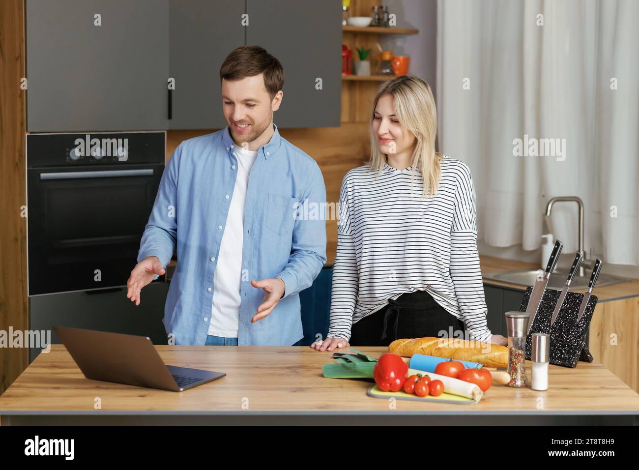 Pleasant family couple standing near big wooden table in modern kitchen ...