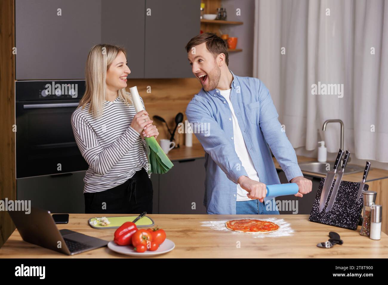 Happy young husband and wife laugh and sing using kitchen appliances ...