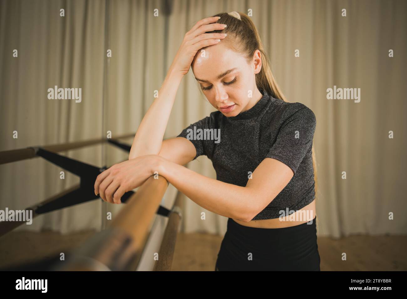 Upset ballerina in training suit stands near ballet barre and looks at ...