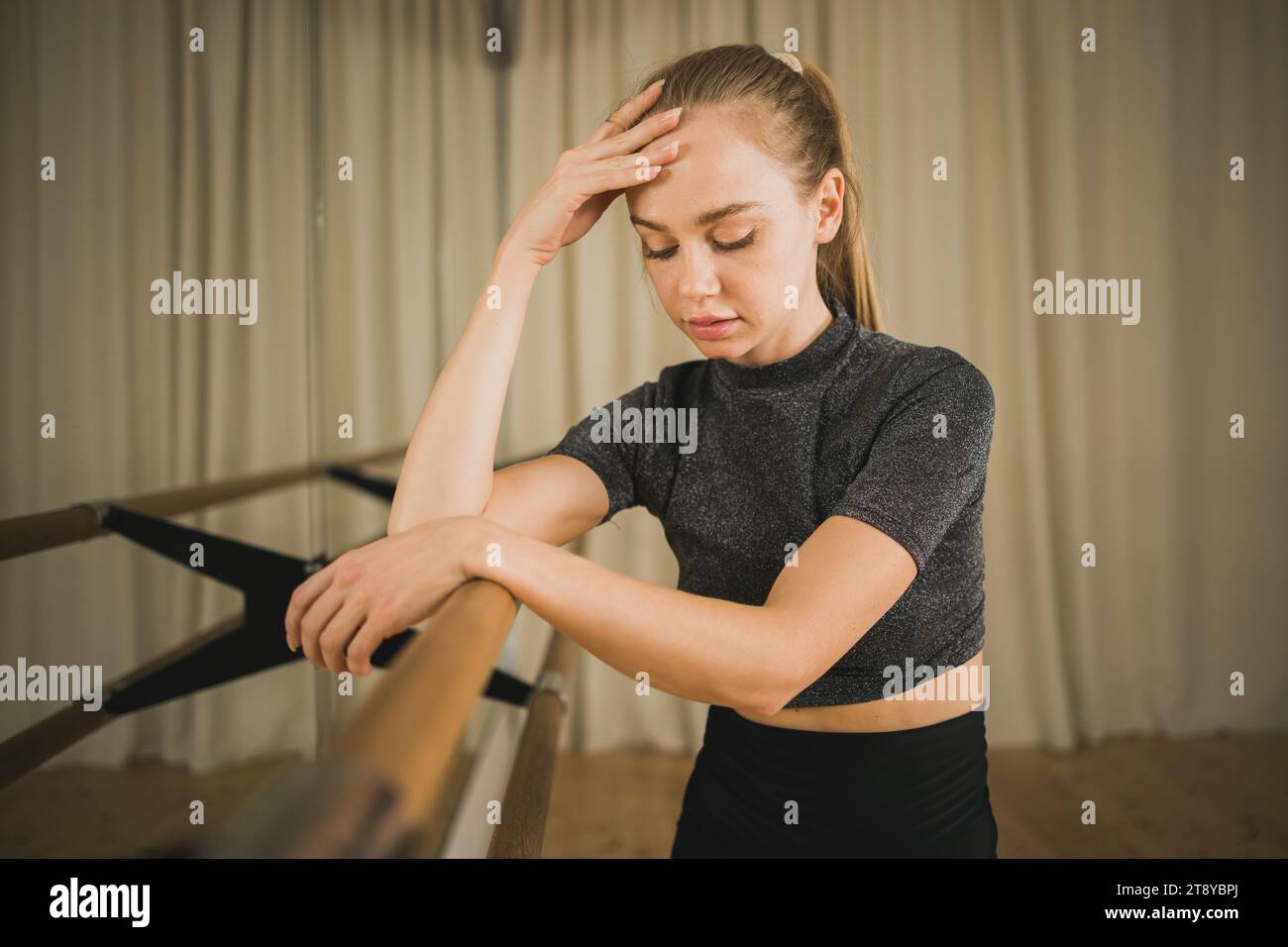 Upset ballerina in training suit stands near ballet barre and looks at ...