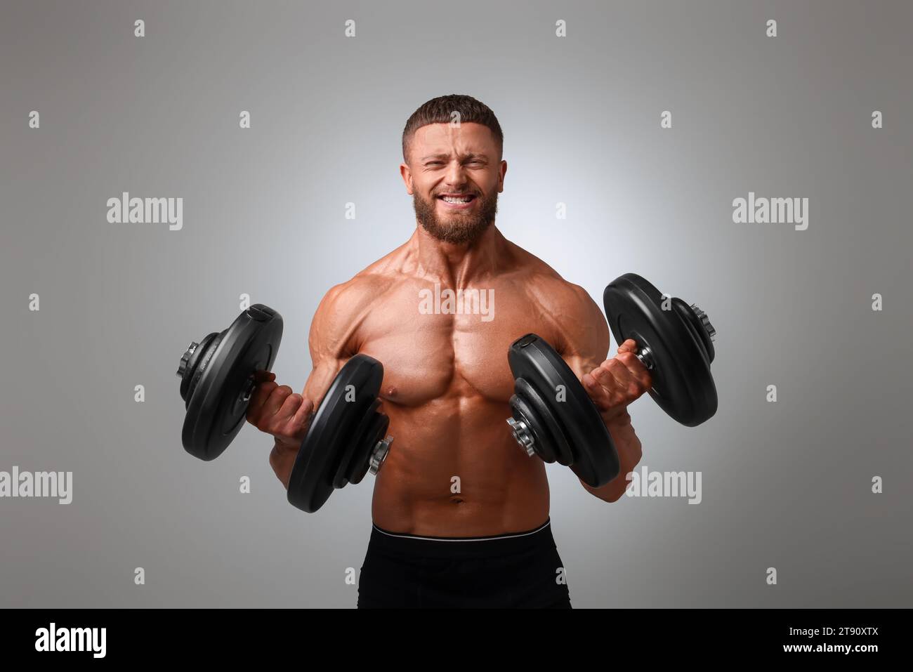 Emotional young bodybuilder exercising with dumbbells on light grey ...