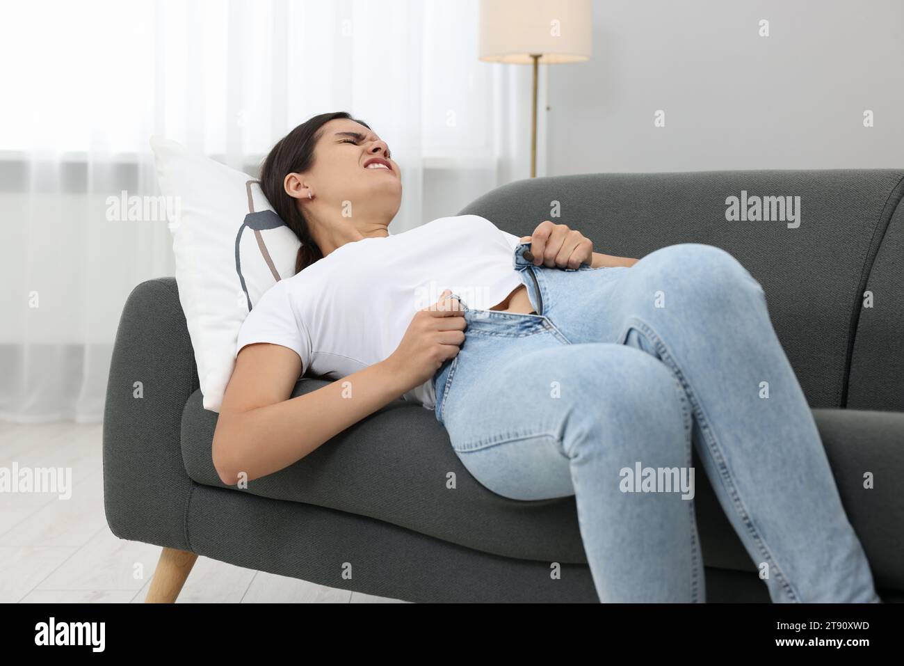 Young woman struggling to squeeze into tight jeans while lying on sofa ...
