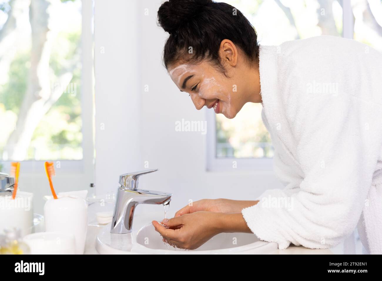 Happy biracial woman in bathrobe rinsing mask from her face in sunny ...
