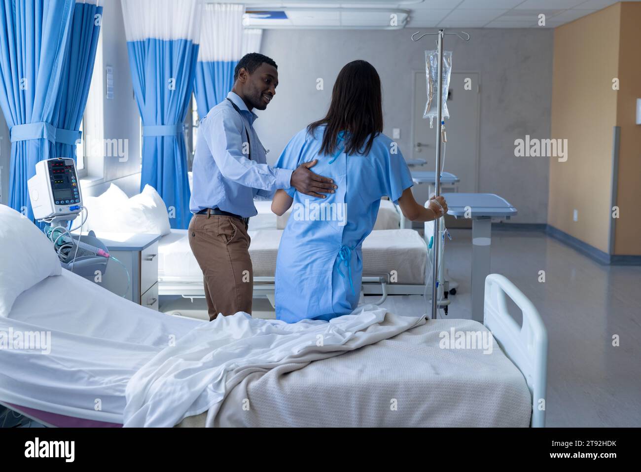 Diverse female patient walking with drip and male doctor helping in ...