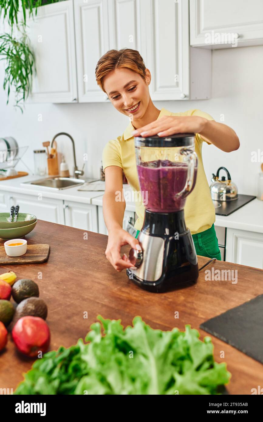delighted vegetarian woman grinding fresh fruits in electric blender ...