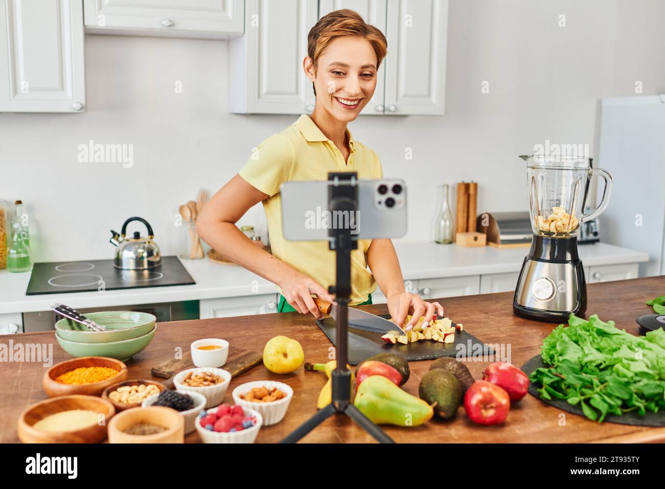 happy vegetarian woman cutting fruits near blender and smartphone on tripod, plant-based diets ...