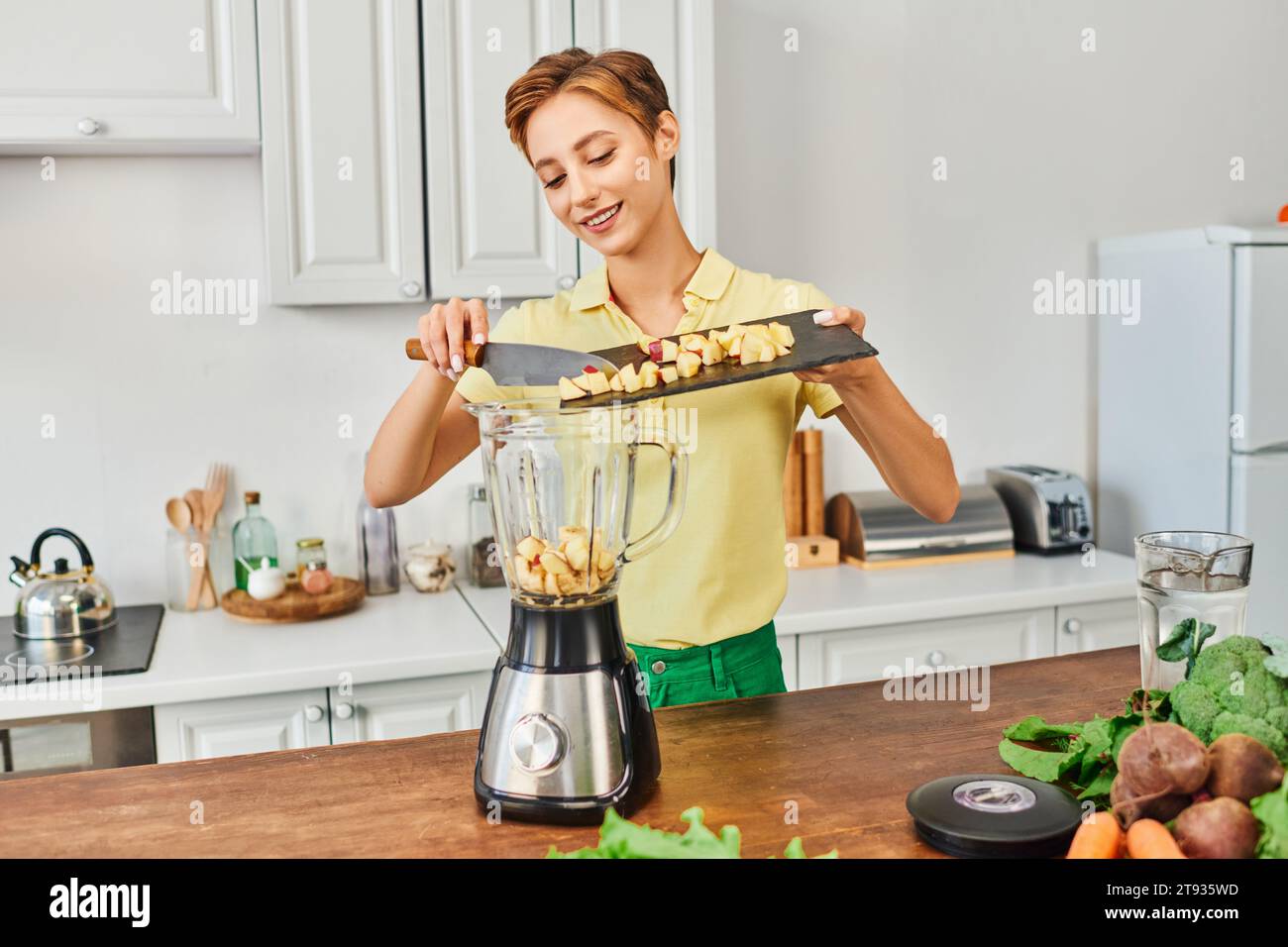smiley woman adding chopped apple in electric blender near plant origin ...