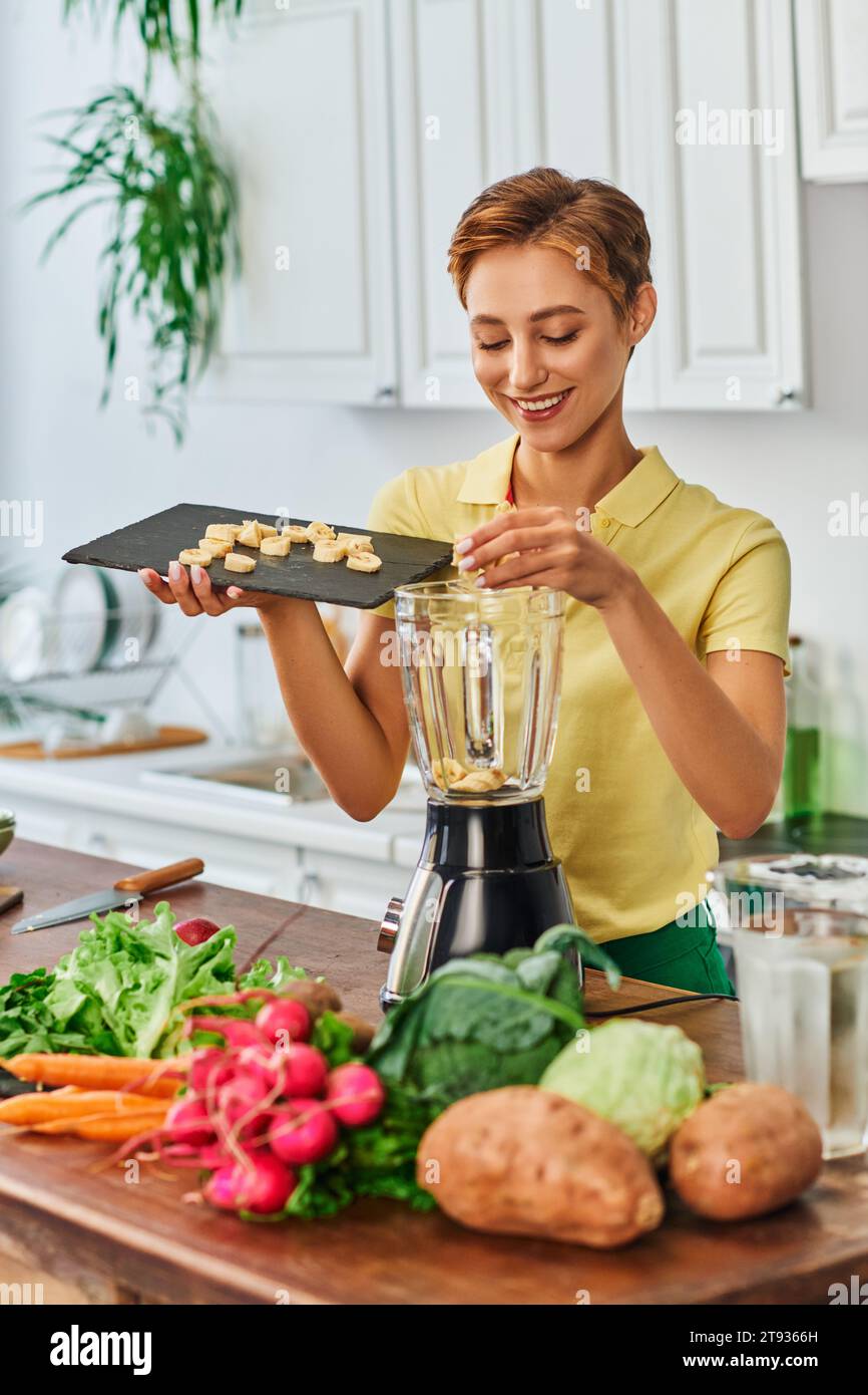 pleased vegetarian woman putting sliced banana in electric blender near ...