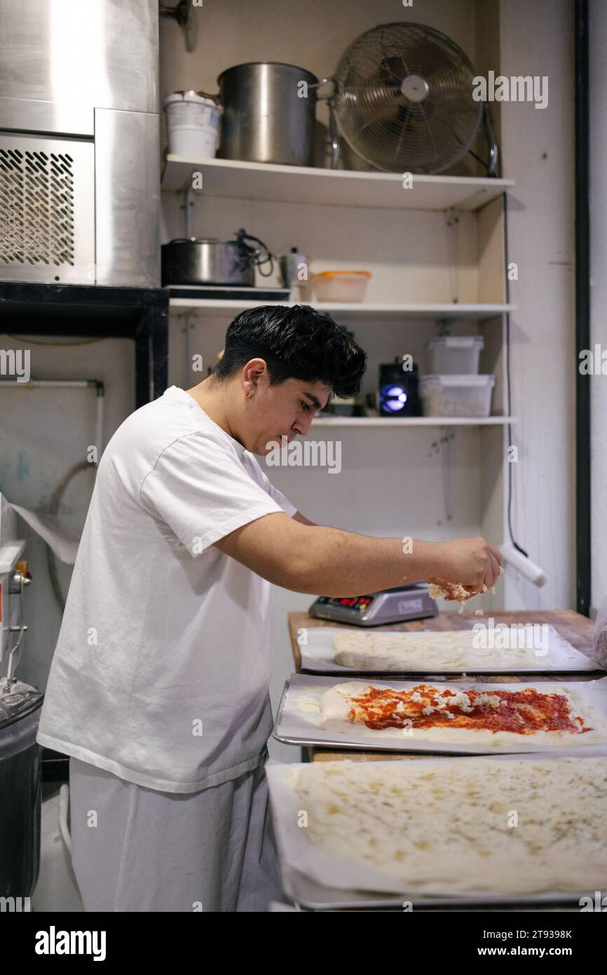 Latino baker dressed in white clothes adding toppings to a pizza in a ...
