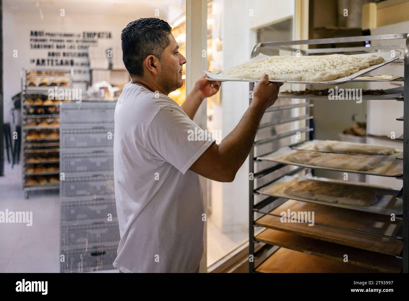 Latino baker dressed in white clothes placing focaccia dough on a tray ...