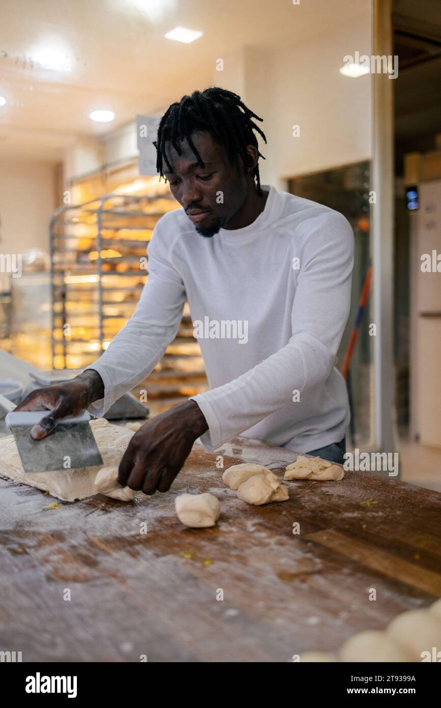 Young black baker with dreadlocks and white clothes cutting dough with ...