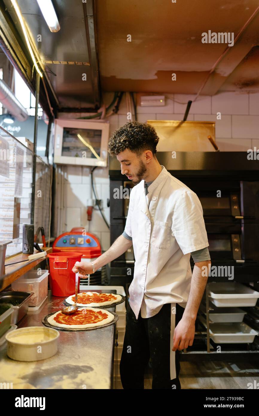 Young arabic pizza man in white apron adding tomato sauce to the base ...