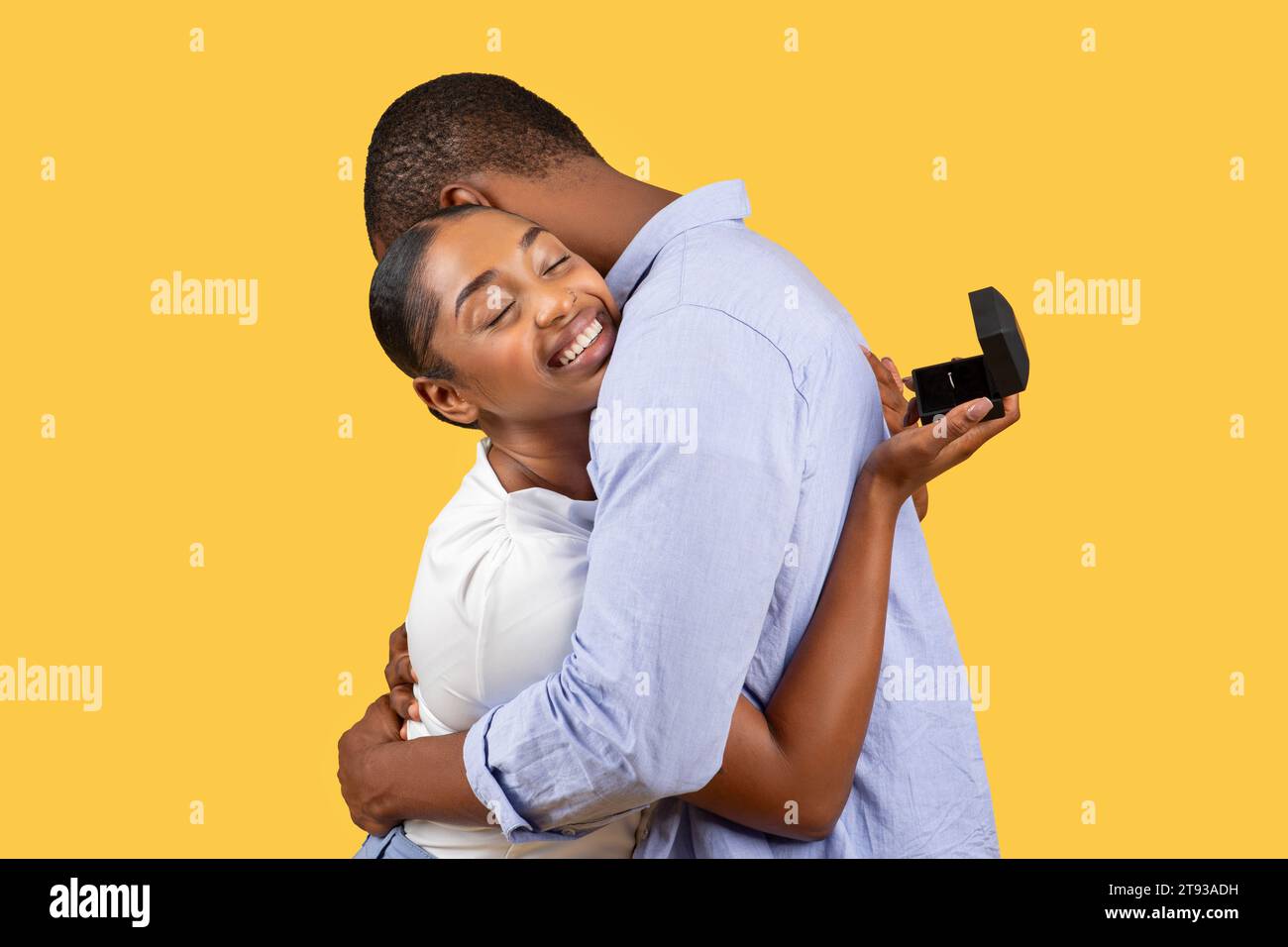 Joyful Black couple hug post-proposal, woman holds ring box, yellow ... Joyful Black couple hug post-proposal, woman holds ring box, yellow ...