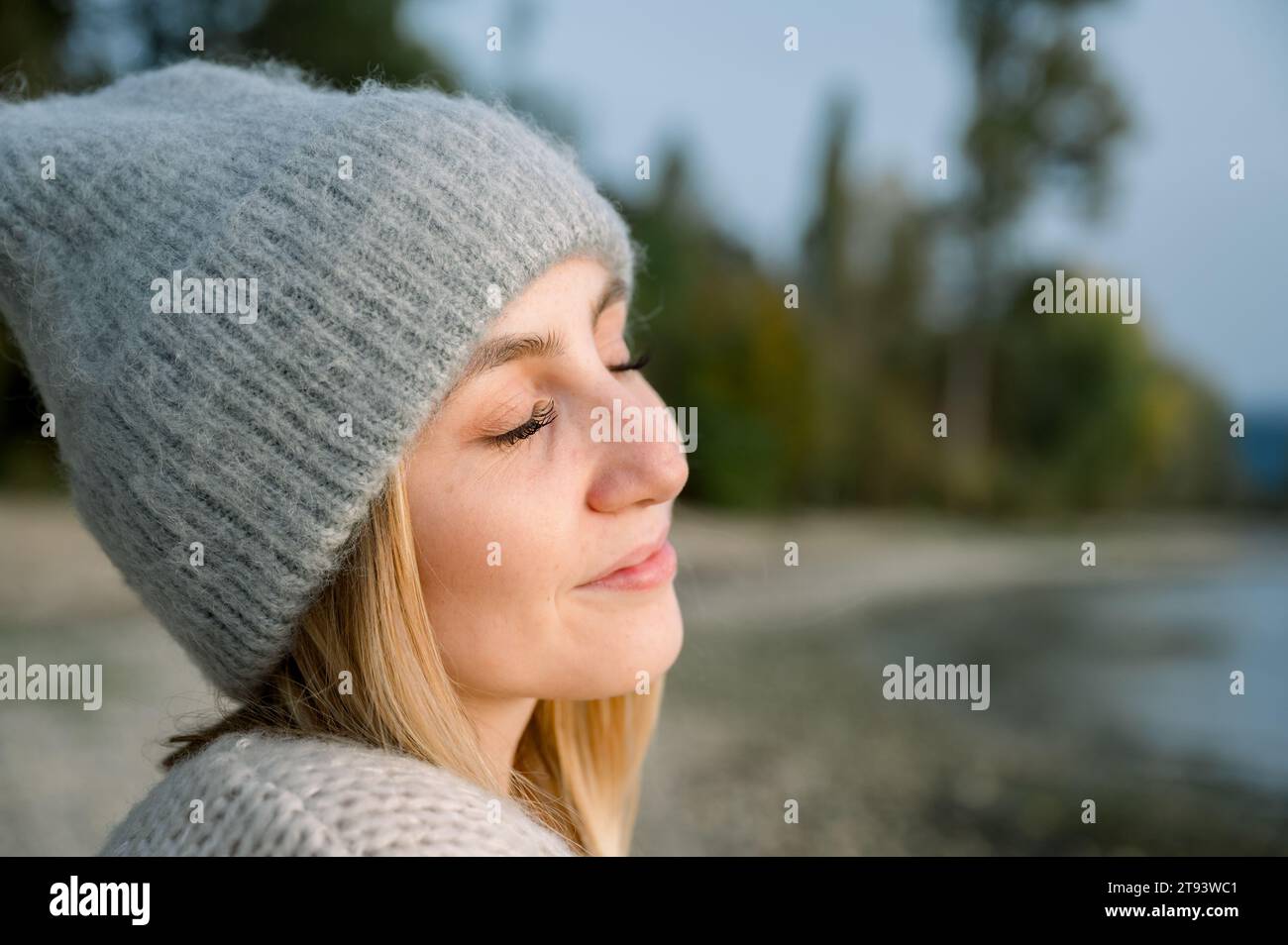 side view of young woman in warm cozy sweater and hat, breathes clean air, looks at the sky with ...