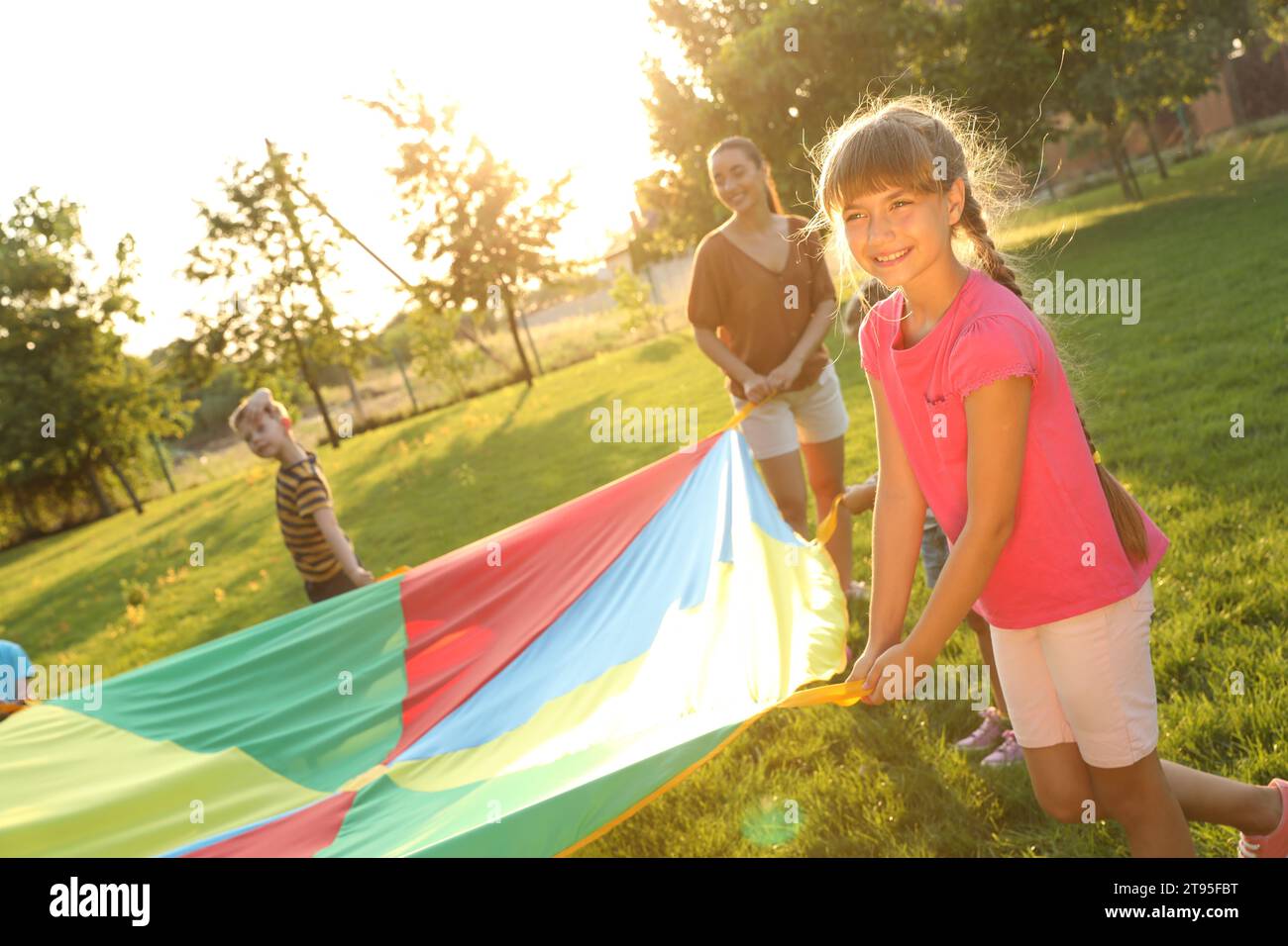 Group of children and teacher playing with rainbow playground parachute on green grass. Summer ...