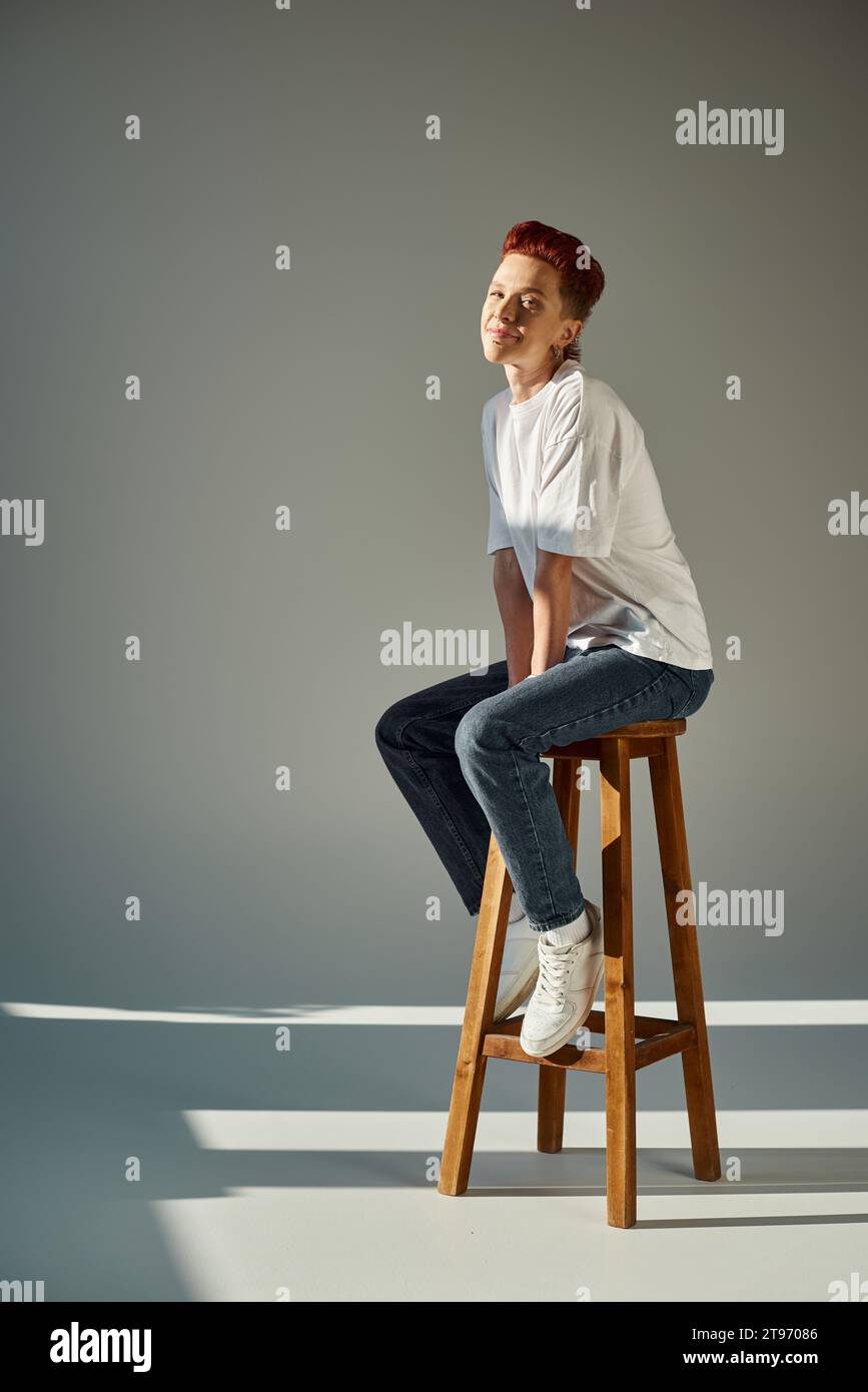 happy non-binary person in white t-shirt and jeans sitting on stool in ...