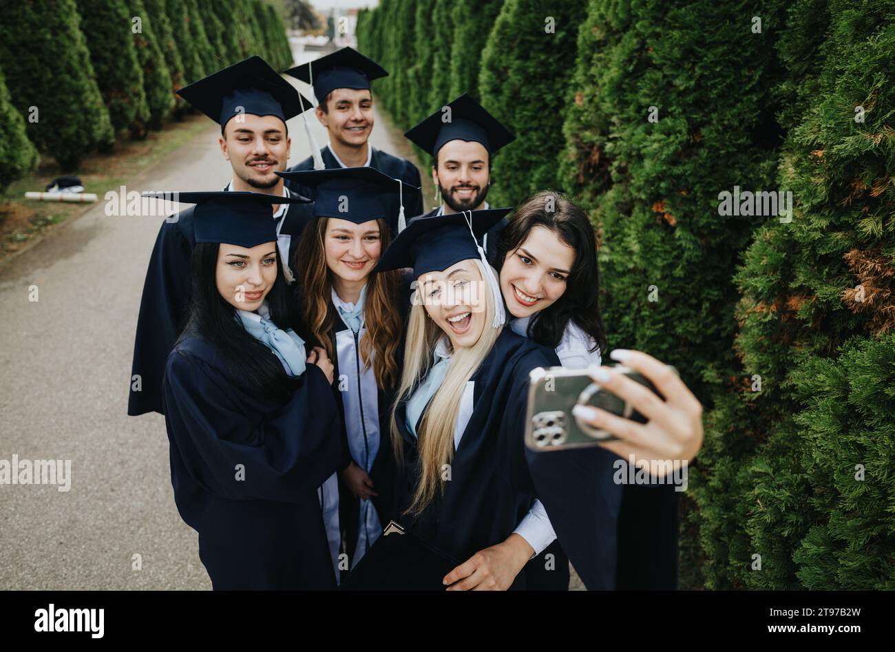 Smiling grads in caps and gowns take a selfie in the park, celebrating ...