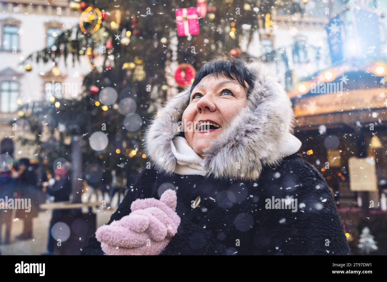 Senior woman in fur hooded coat looking up joyfully as snow falls at ...