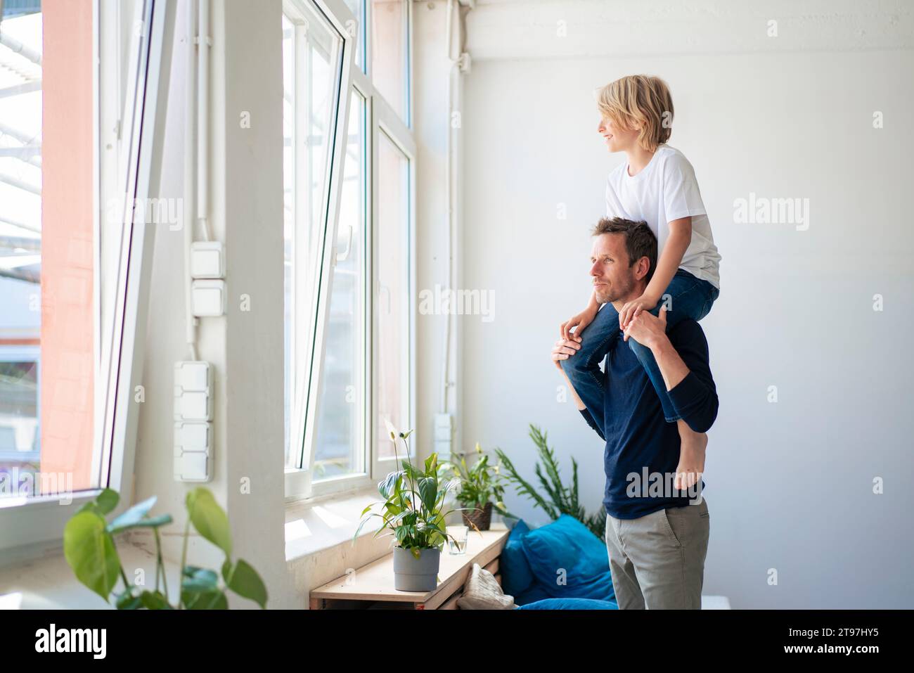 Thoughtful father carrying son on shoulders and looking through window ...