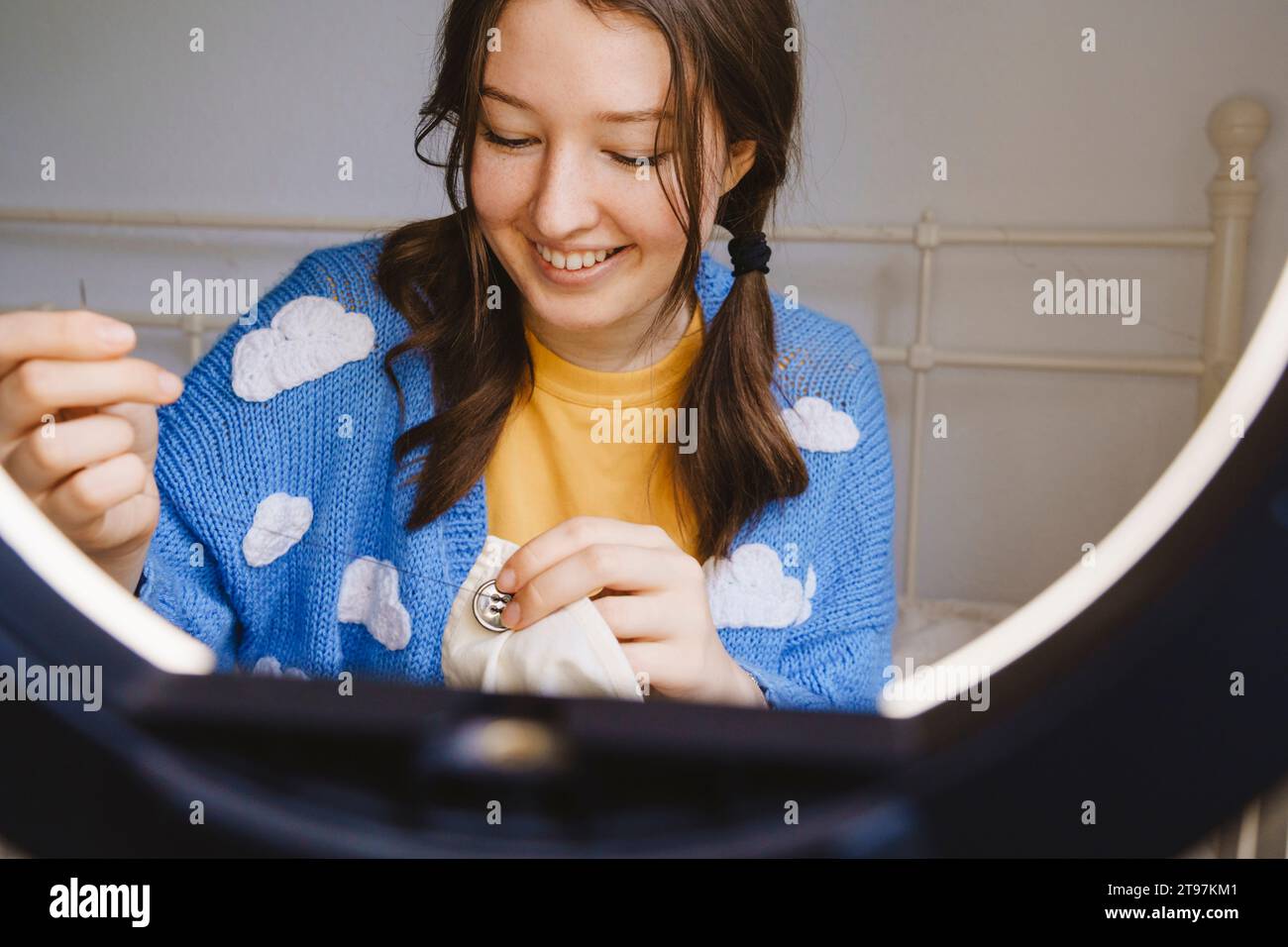 Smiling teenage girl sewing button in front of ring light Stock Photo ...
