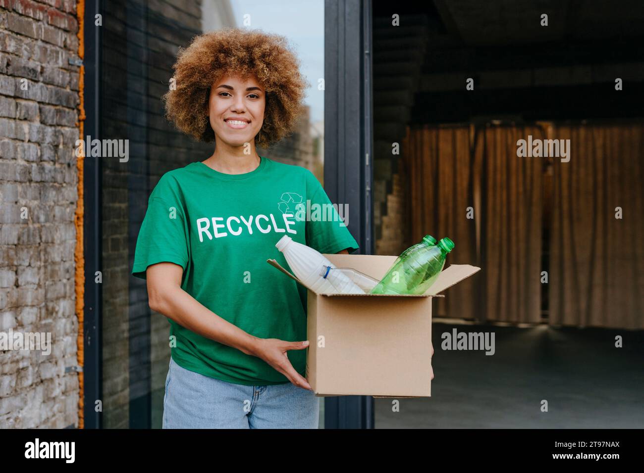 Smiling activist holding cardboard box with plastic bottles in front of ...