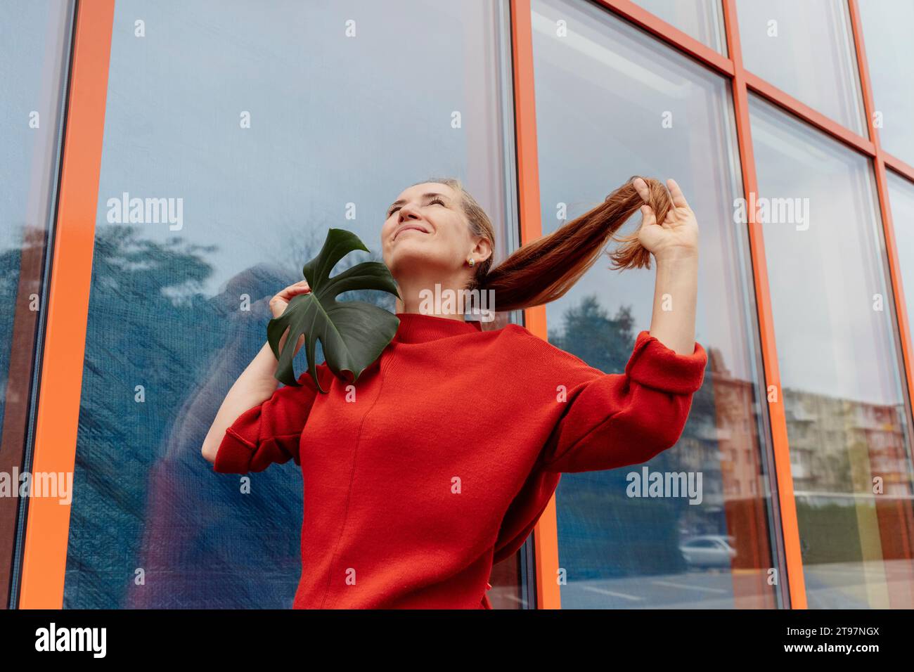 Smiling woman holding monstera leaf and pulling ponytail in front of ...