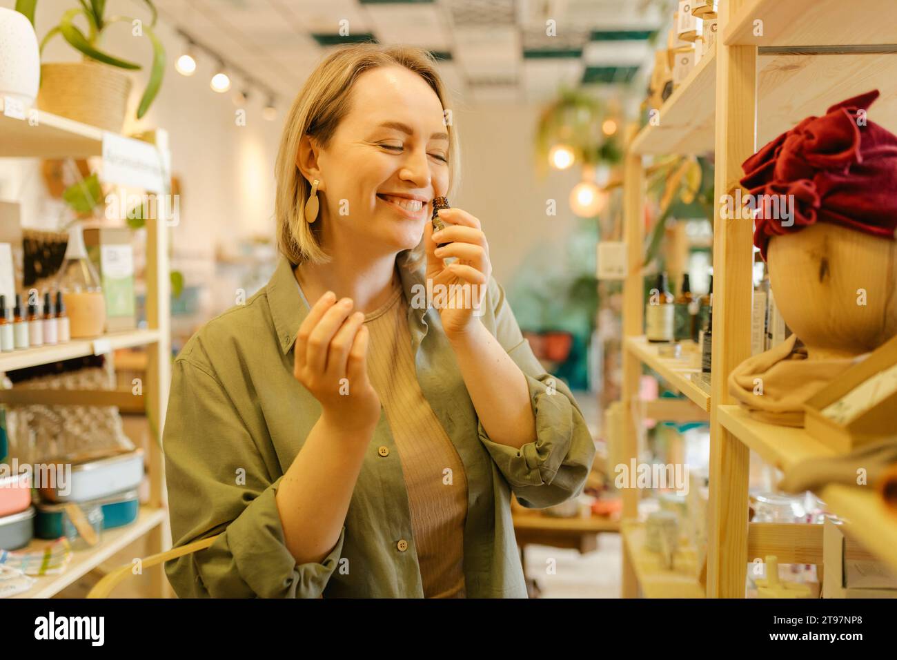 Smiling woman with eyes closed smelling face oil in zero waste store ...