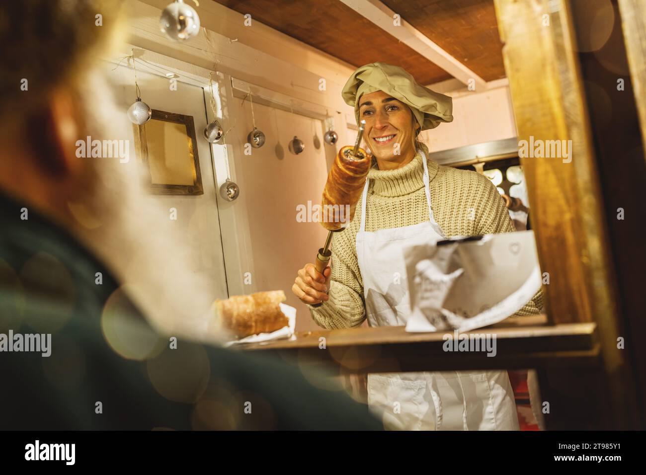 Happy saleswoman shows an old man a finshed Baumstriezel, trdelnik or ...