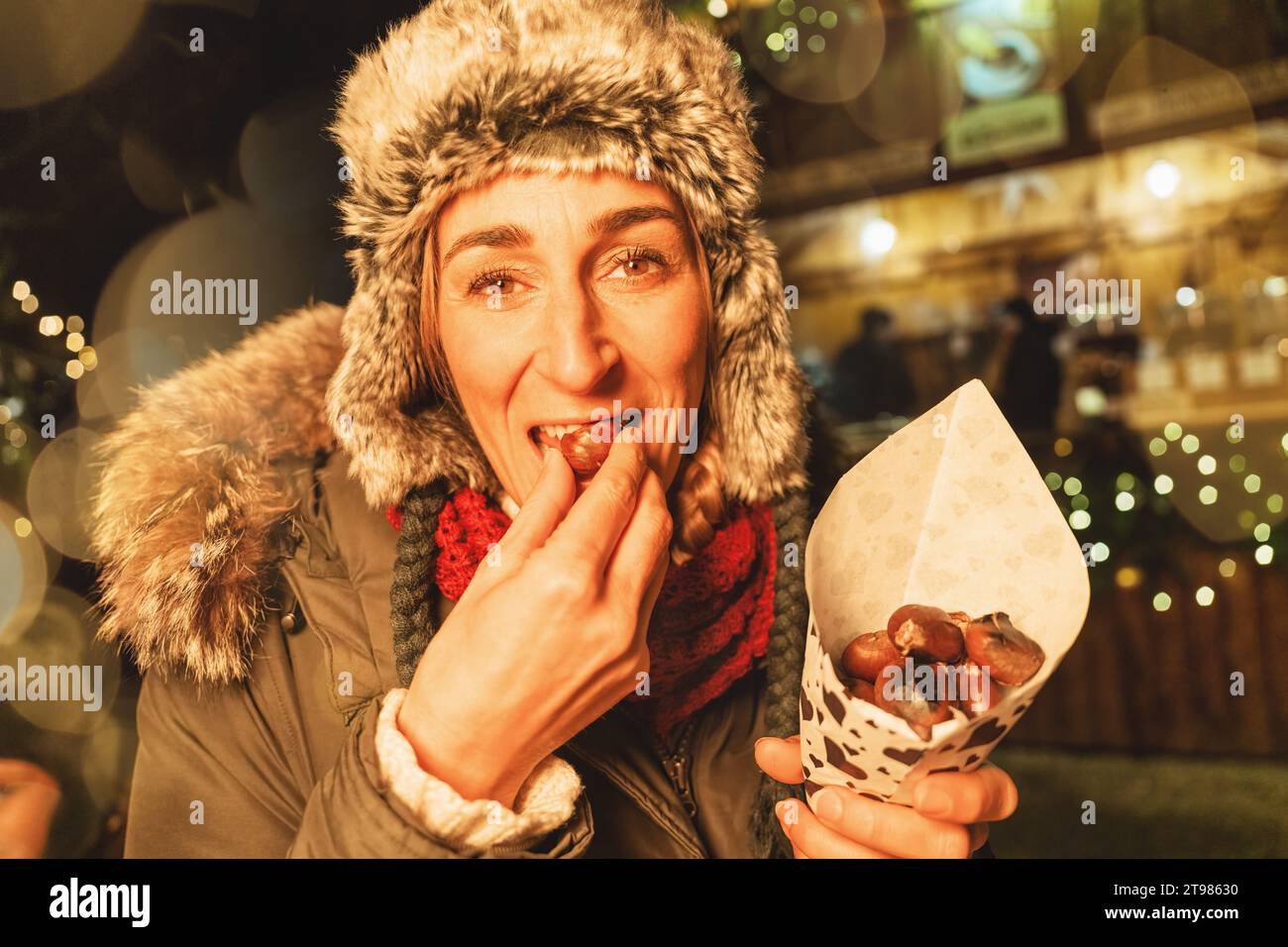 Woman eating roasted chestnuts at a Christmas market, delighted, with festive lights around ...