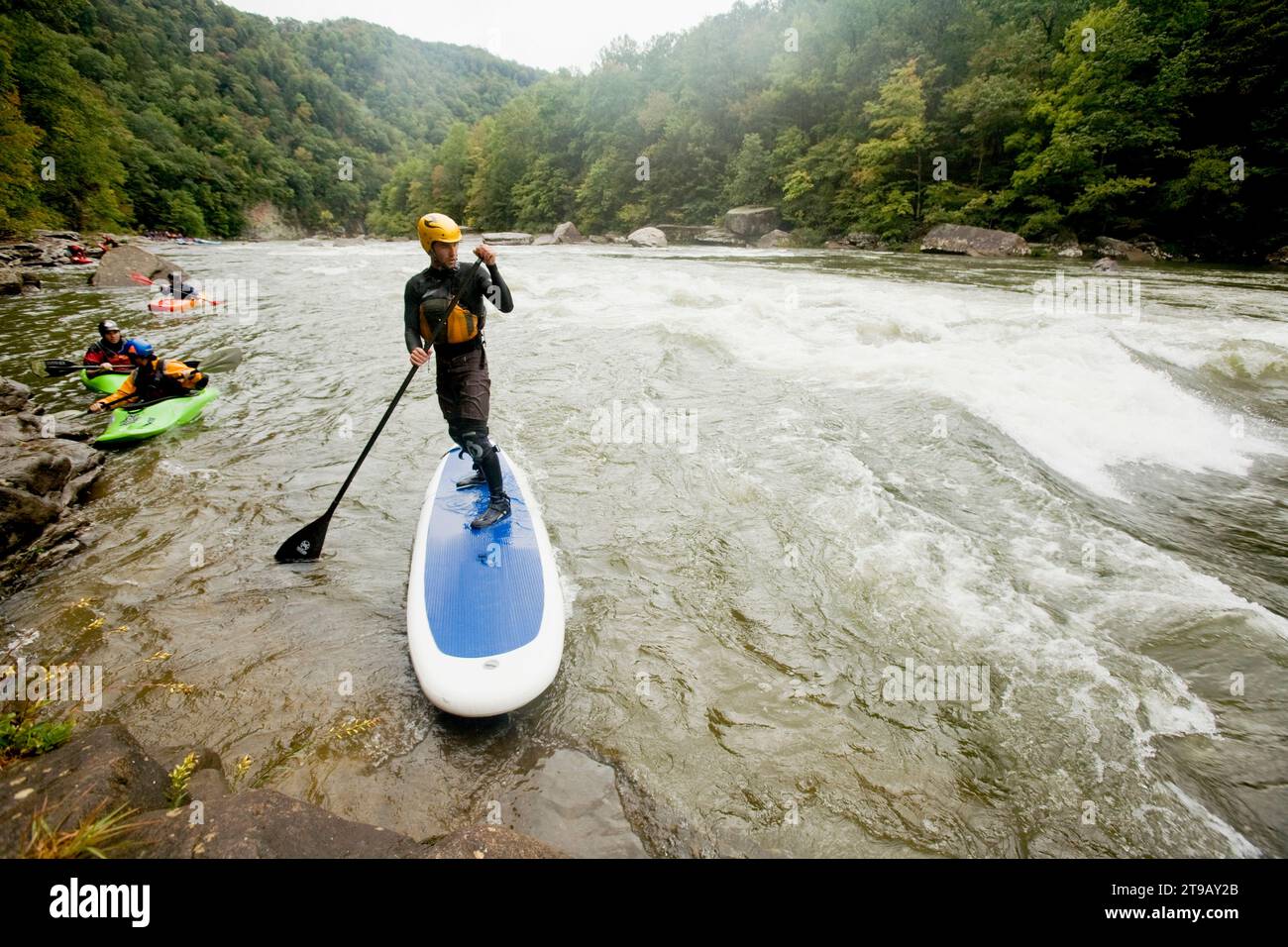 A stand up paddleboarder paddling up to a wave on a river with kayakers ...