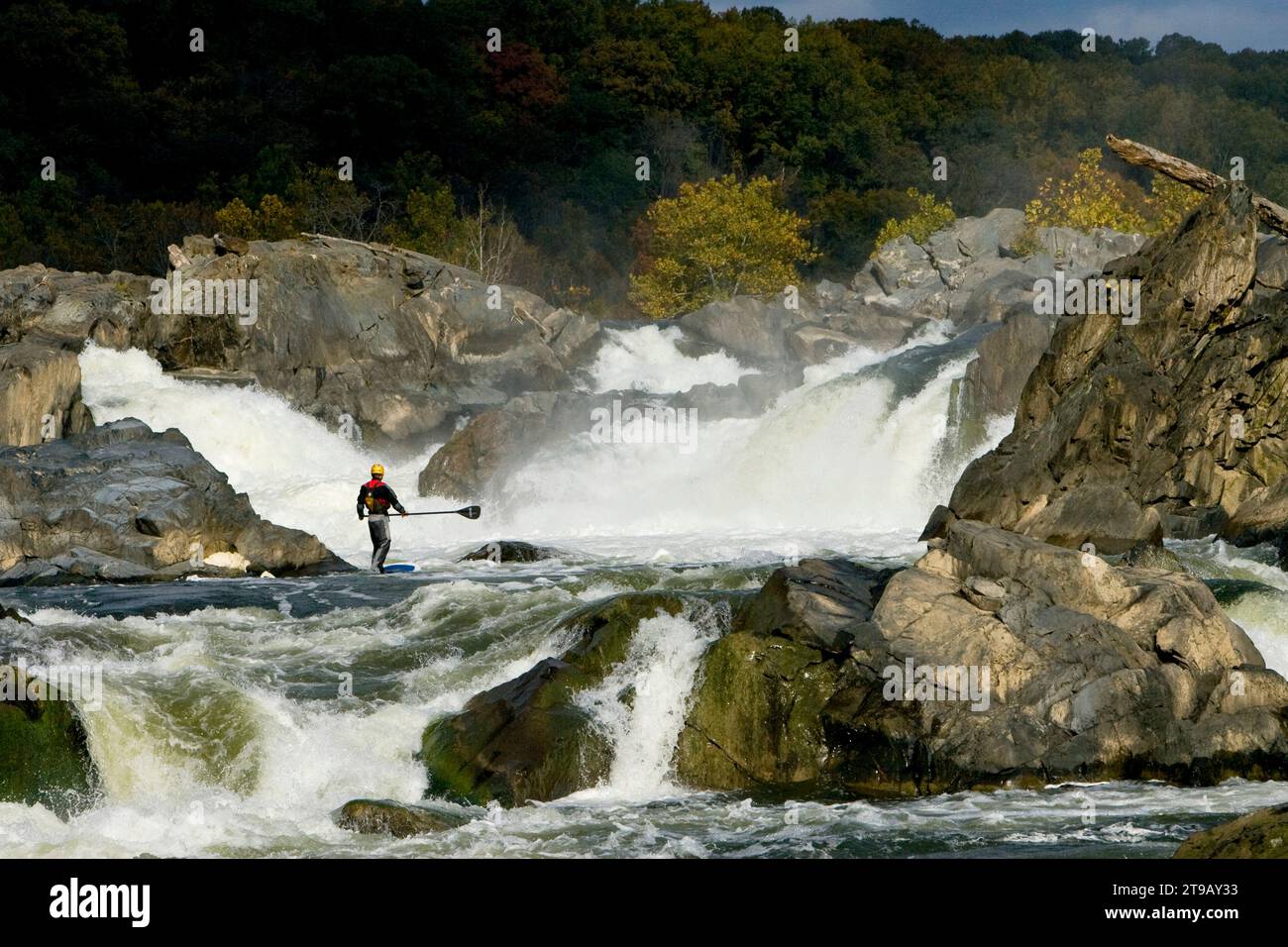 One man stand up paddleboarding at the base of a big set of waterfalls ...