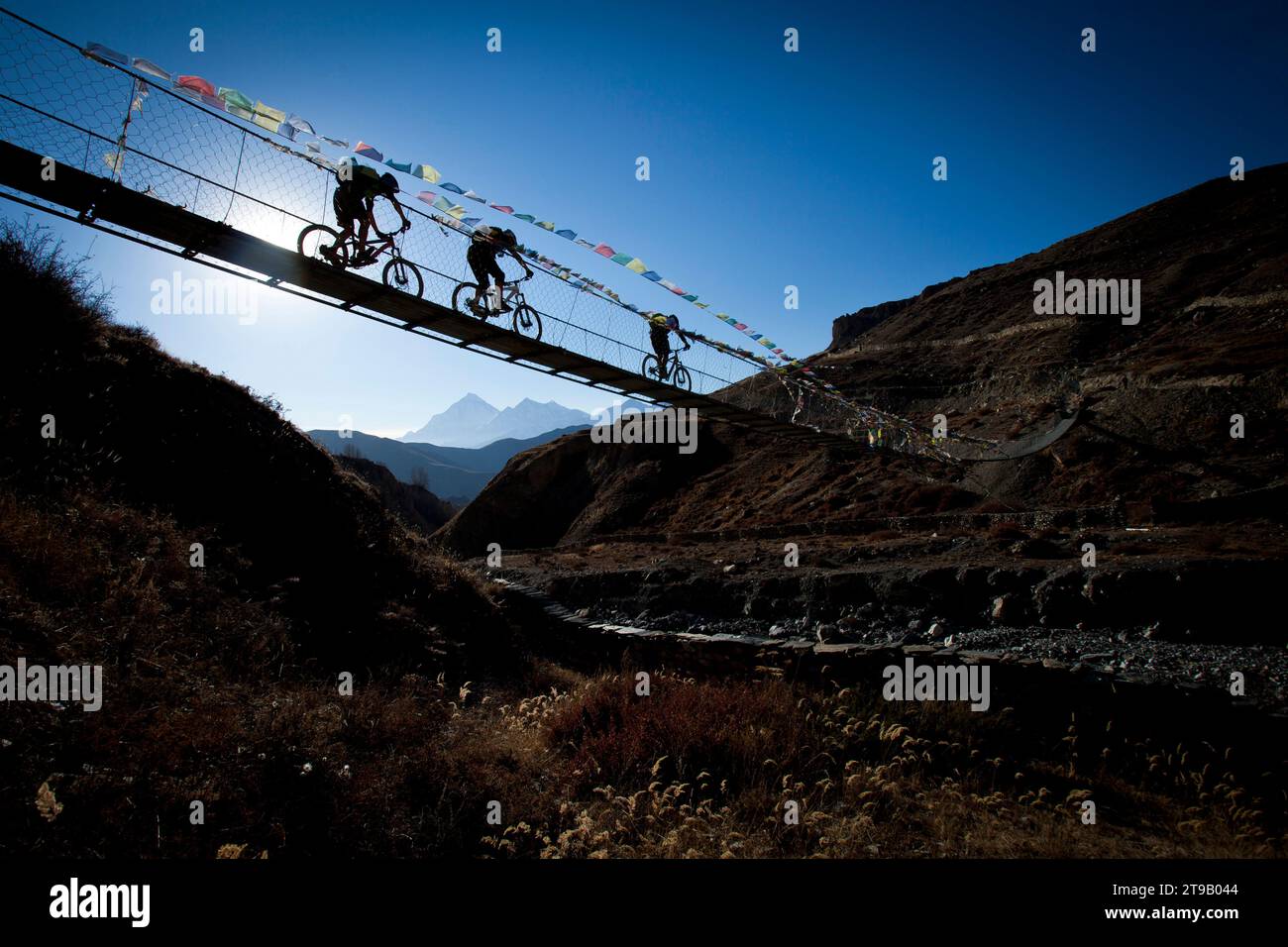 Three men ride mountain bikes across a small bridge with prayer flags ...