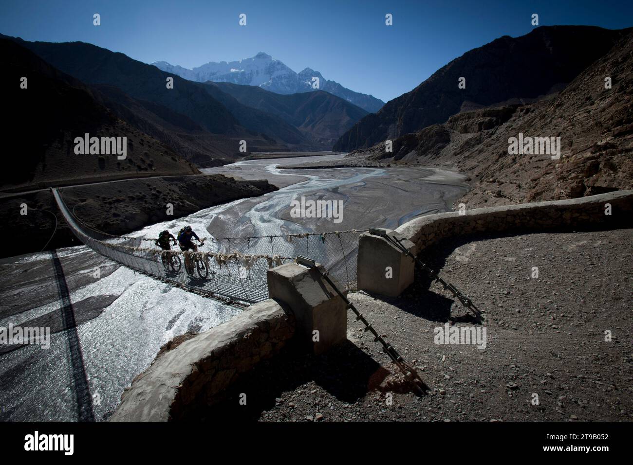 Two men ride mountain bikes across small bridge over river valley with ...
