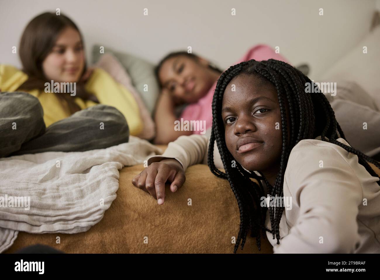 Teenage girl with braided hair by female friends lying on bed in ...