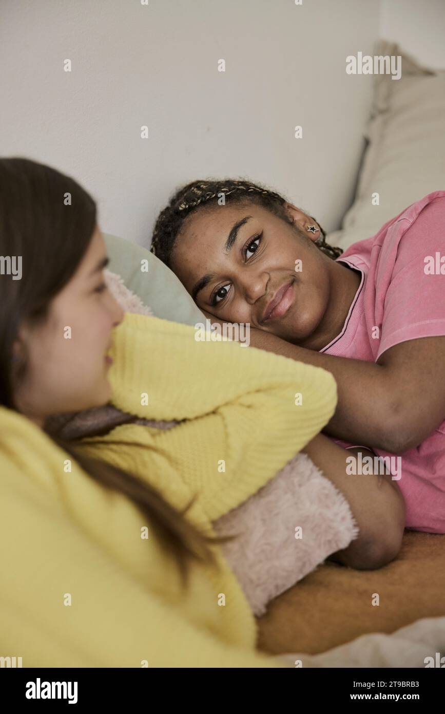 Smiling teenage girl resting on bed with female friend in bedroom at ...