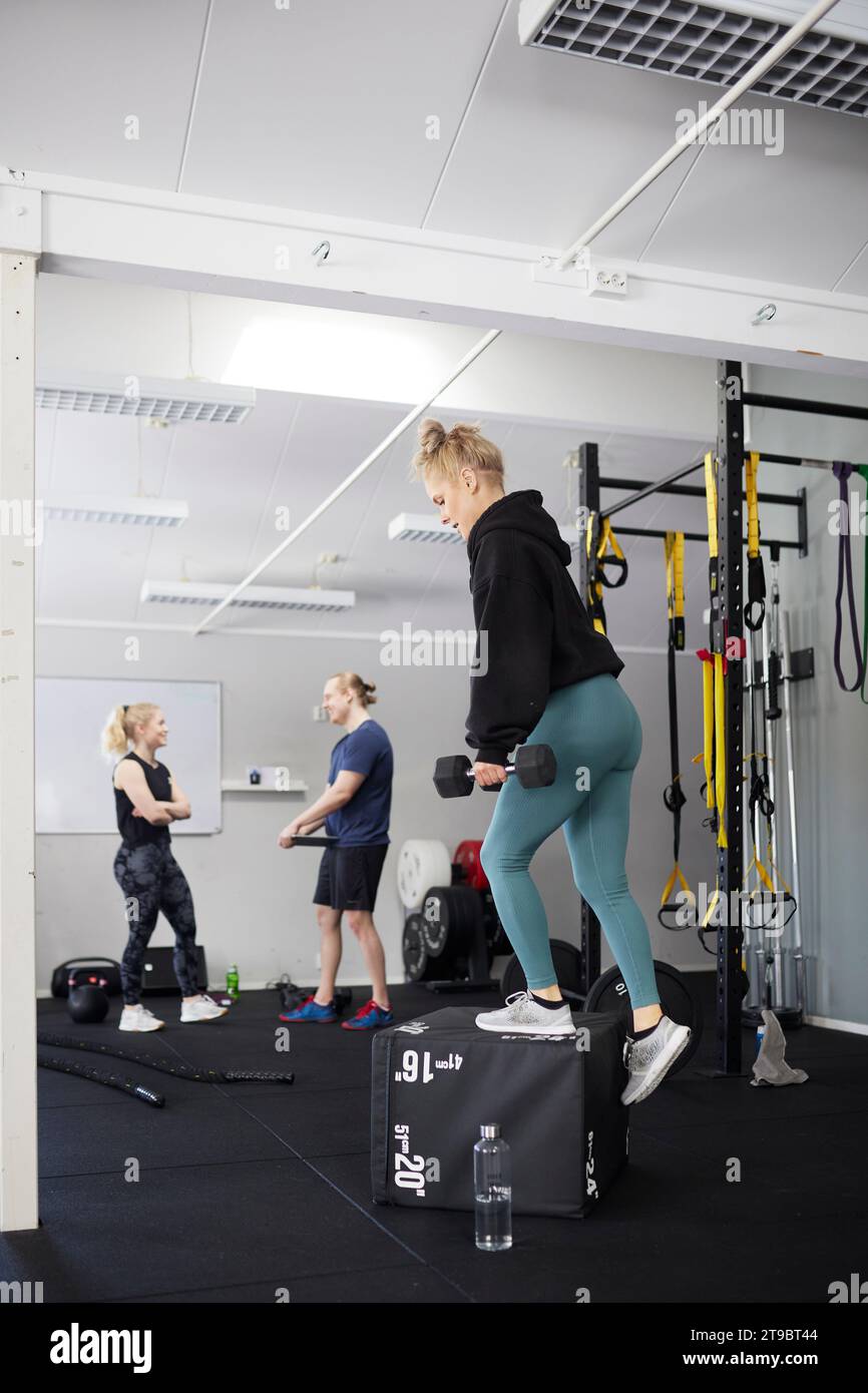 Full length side view of young woman with dumbbell exercising on block ...