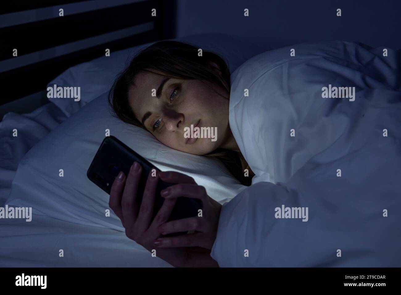 A girl in bed with a phone on a white bed in a dark room Stock Photo ...