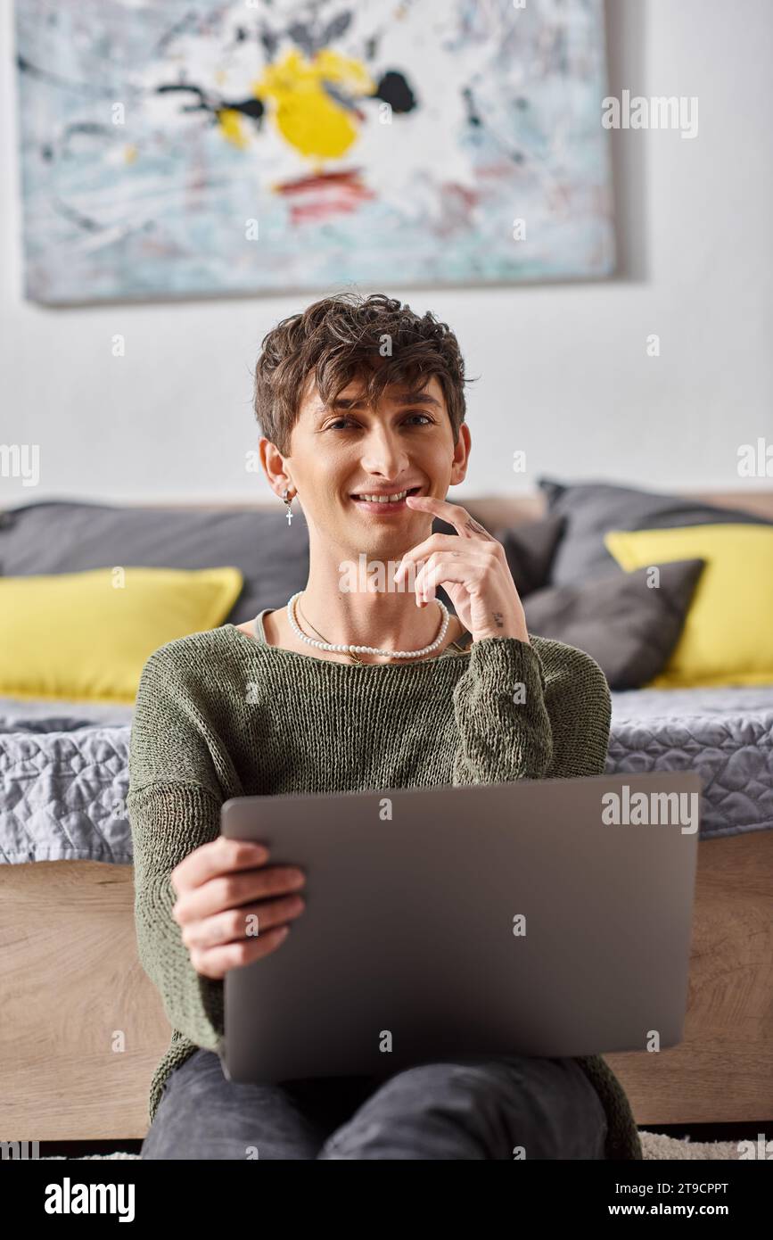pleased and curly transgender influencer using laptop and sitting next ...