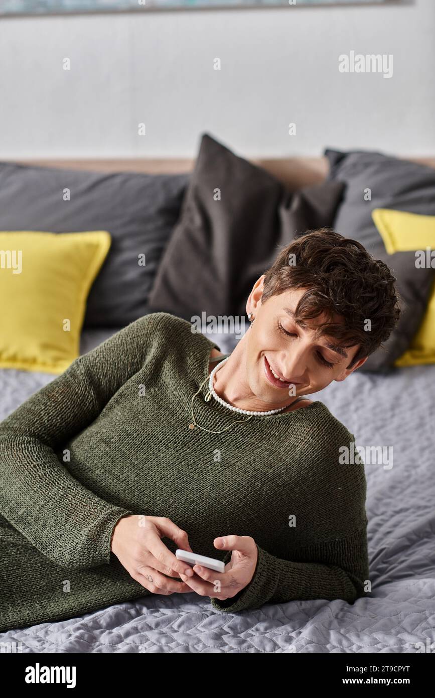 cheerful and curly transgender blogger using smartphone and lying on ...