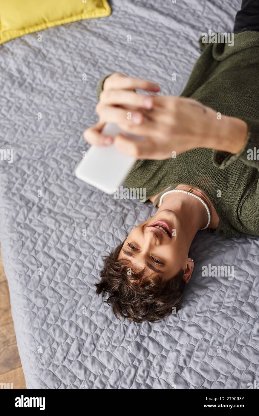 happy transgender blogger with curly hair using smartphone and lying on ...