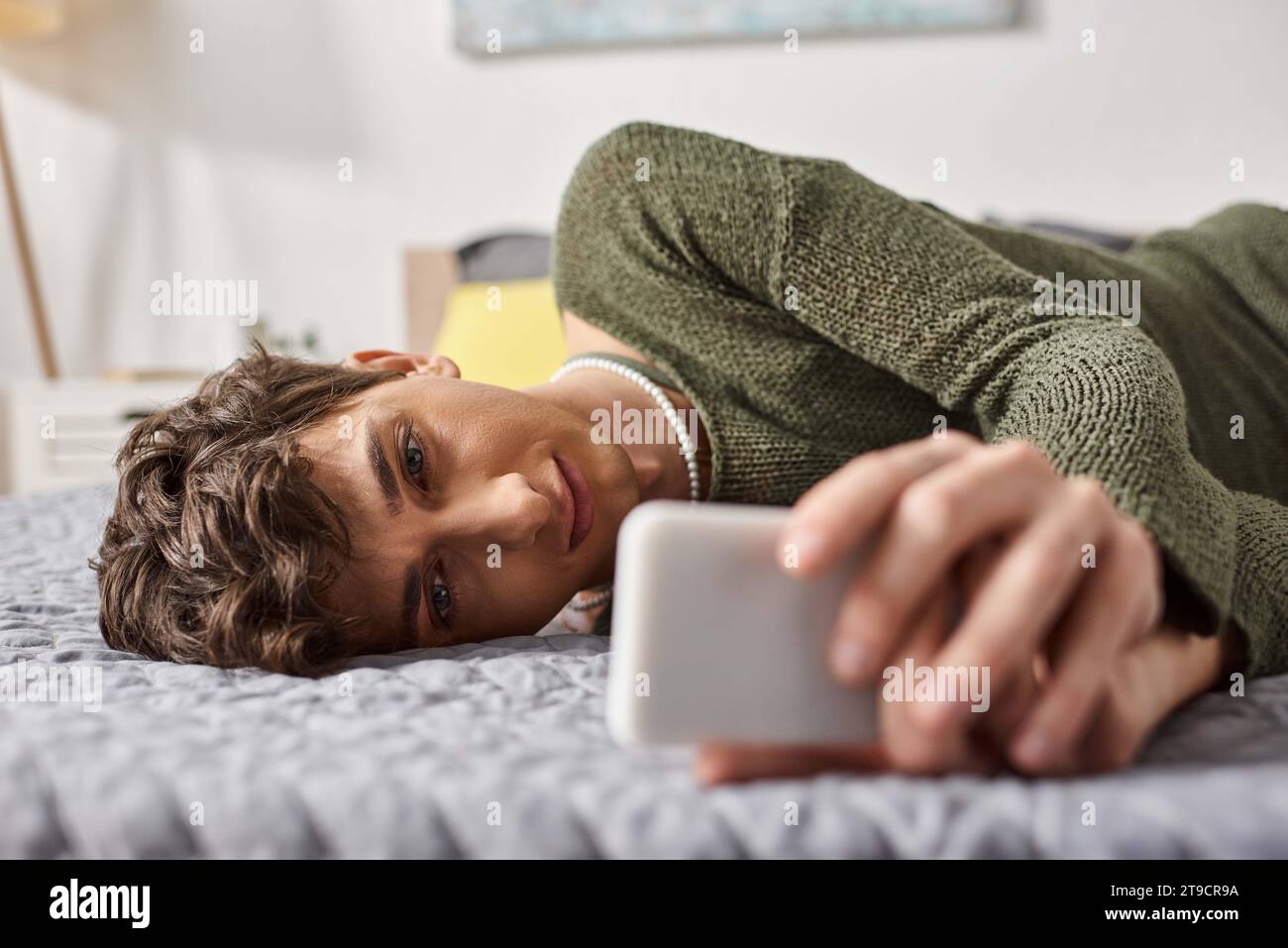 transgender blogger with curly hair using smartphone and lying on bed ...